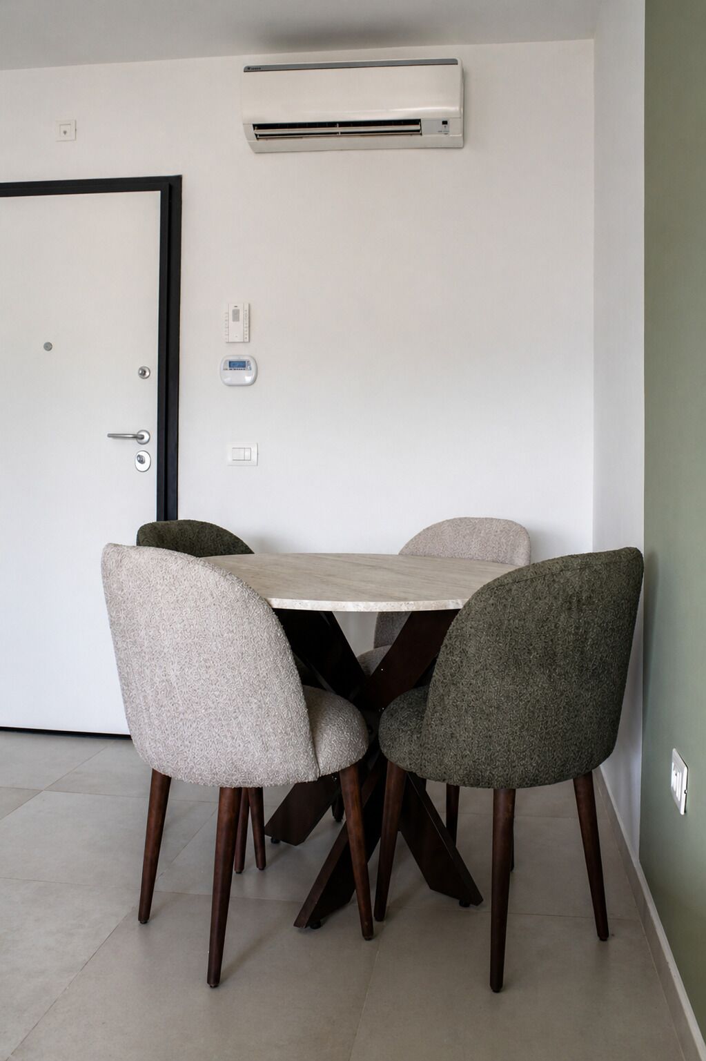 Uma área de refeições com mesa e cadeiras, um aparelho de ar condicionado acima, uma porta com puxador e um termostato na parede.