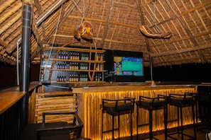 Uma área de bar com móveis de madeira, uma estante com garrafas e uma tela de televisão.