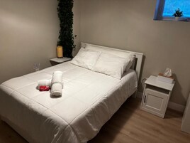 Uma cama bem arrumada com lençóis brancos, uma pequena mesa de cabeceira com um estojo de lenços e uma janela com vista para o verde.