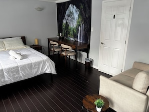 Quarto de hotel com cama, escrivaninha com cadeiras, pequena mesa com planta, porta branca e parede com mural de cachoeira.