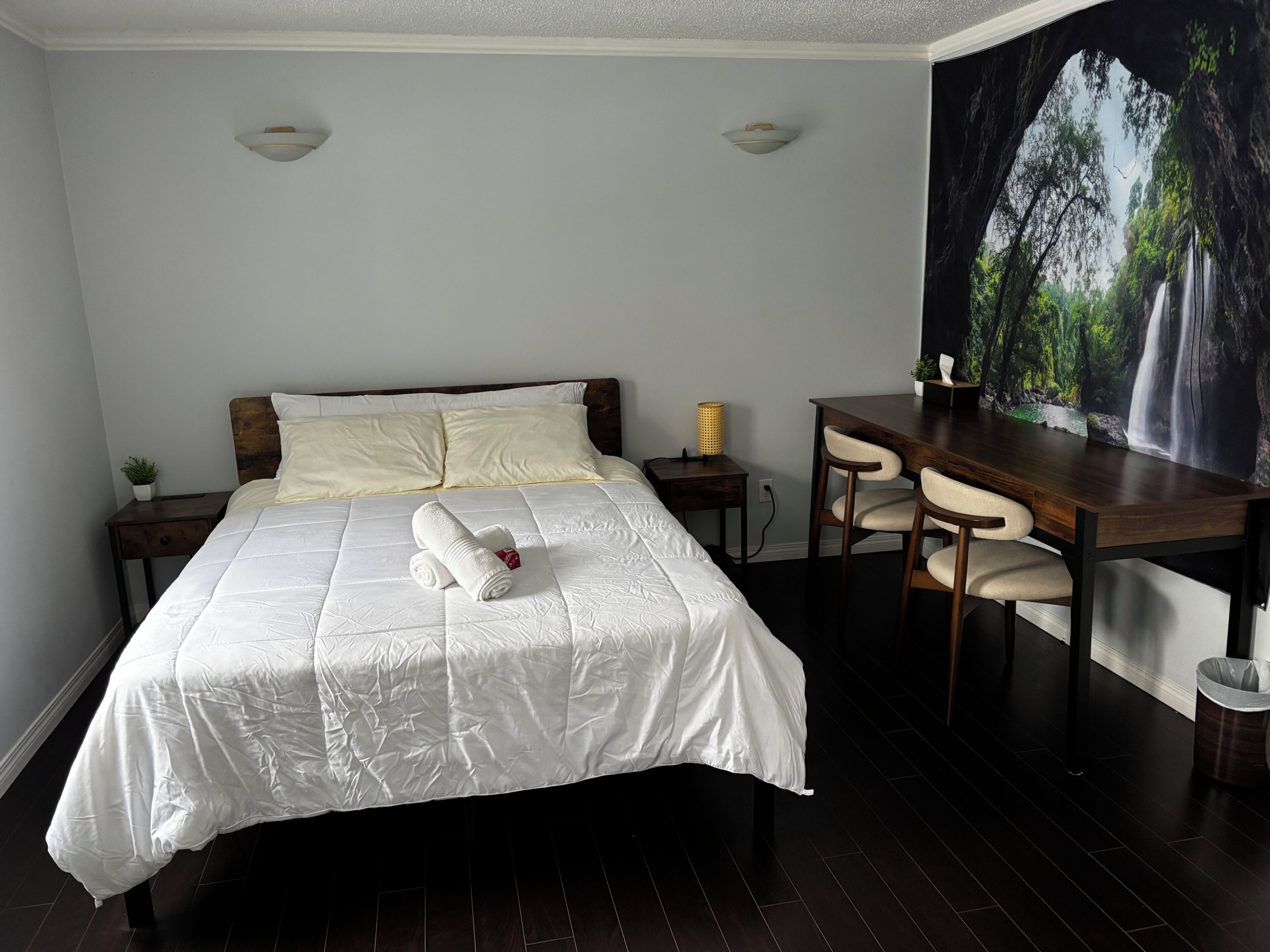 Quarto com cama, escrivaninha, duas cadeiras, uma pequena planta e um quadro de uma cascata.