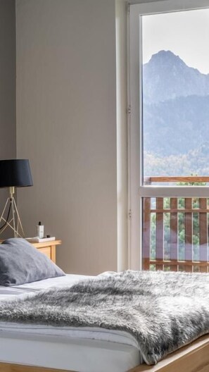 Quarto com cama, mesa de cabeceira, luminária e vista para montanhas pela janela.