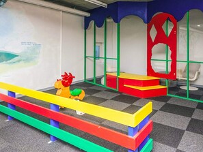Uma área de recreação colorida com piso xadrez, um brinquedo vermelho e verde, e um banco amarelo e vermelho.