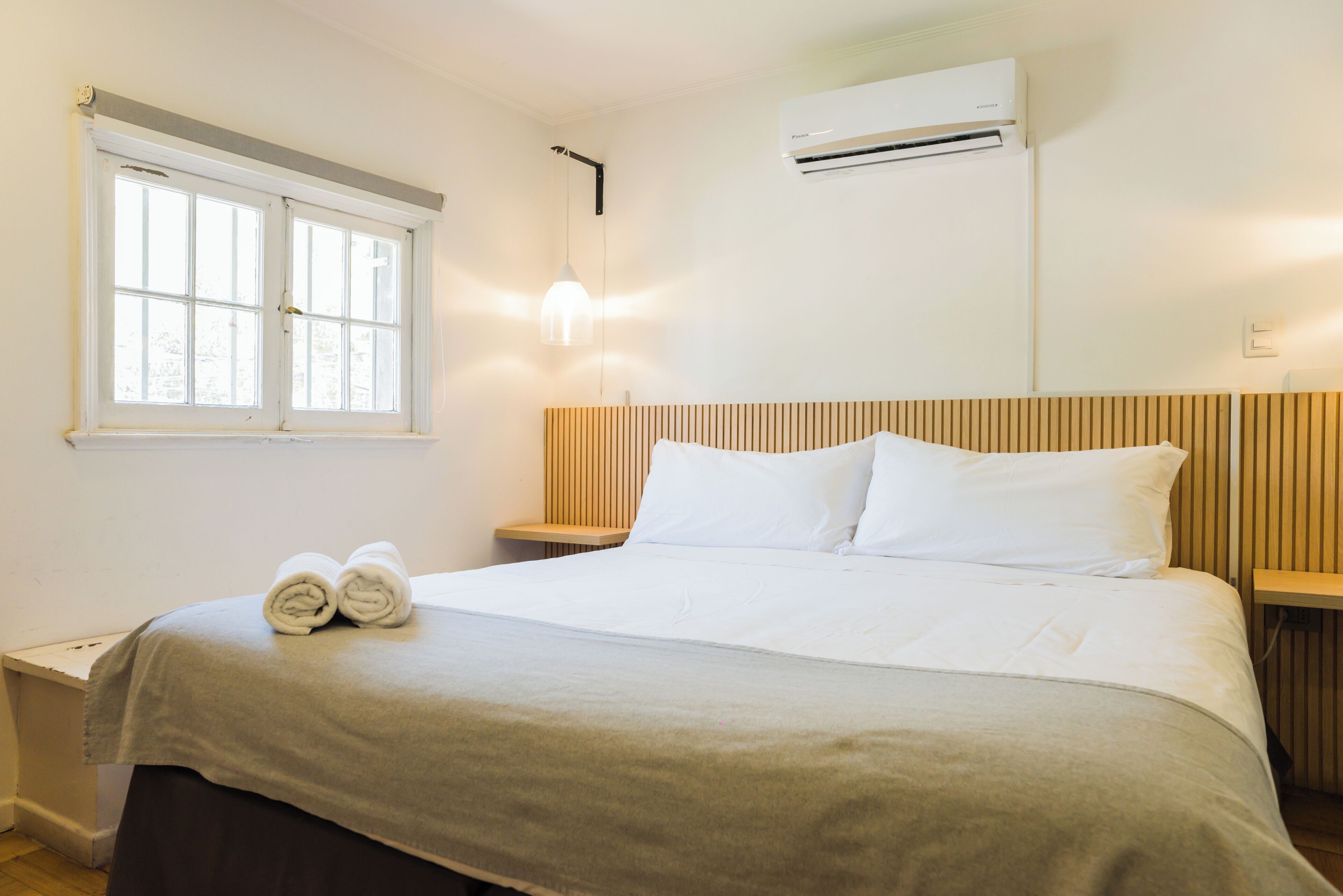 Uma cama bem arrumada com lençóis brancos, uma janela com persianas, um ar condicionado fixado na parede e uma mesa de cabeceira com abajur.