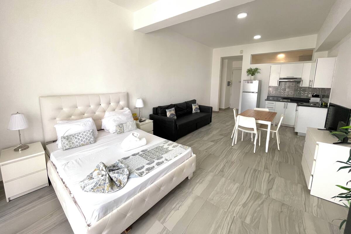 Quarto de hotel moderno com cama, mesa de cabeceira, luminária e um espaço com kitchenette.