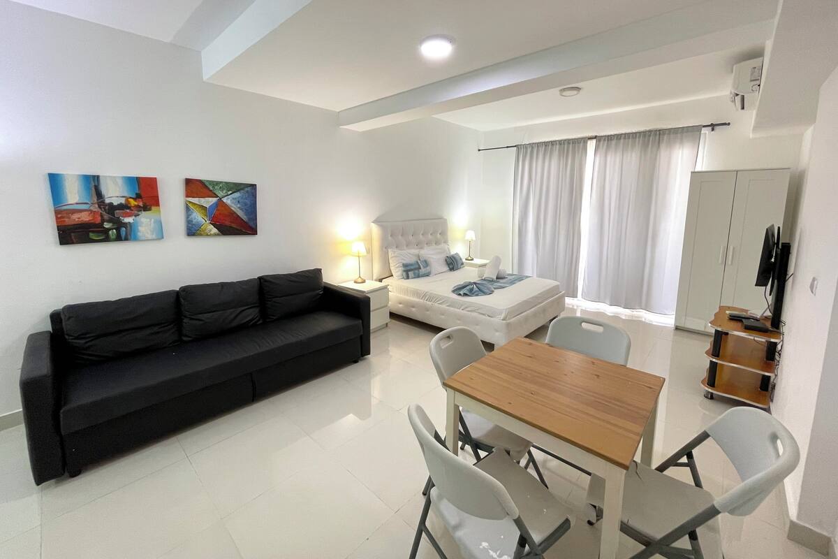 Quarto de hotel moderno com cama, sofá preto, mesa de jantar com cadeiras, televisão e guarda-roupa.
