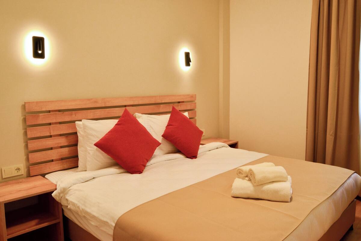 Cama bem arrumada com cabeceira de madeira, dois travesseiros vermelhos e uma toalha branca dobrada ao lado.