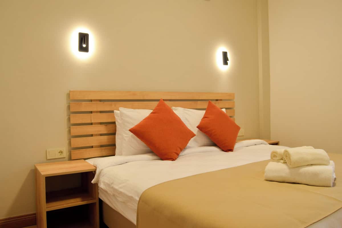 Quarto de hotel com cabeceira de madeira, duas almofadas laranja, colcha branca e um par de toalhas enroladas na cama.
