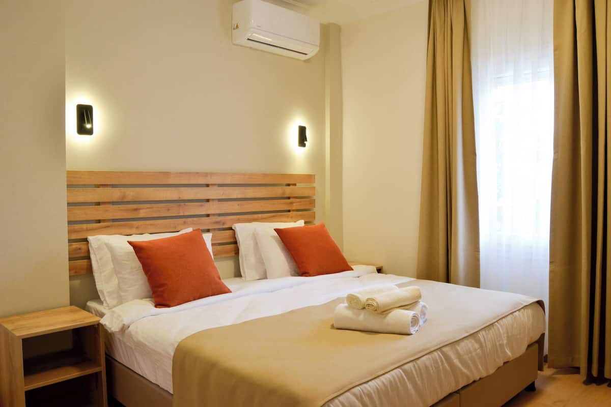 Quarto de hotel com cabeceira de madeira, duas almofadas, cama com roupa de cama branca, mesa de cabeceira e aparelho de ar condicionado.