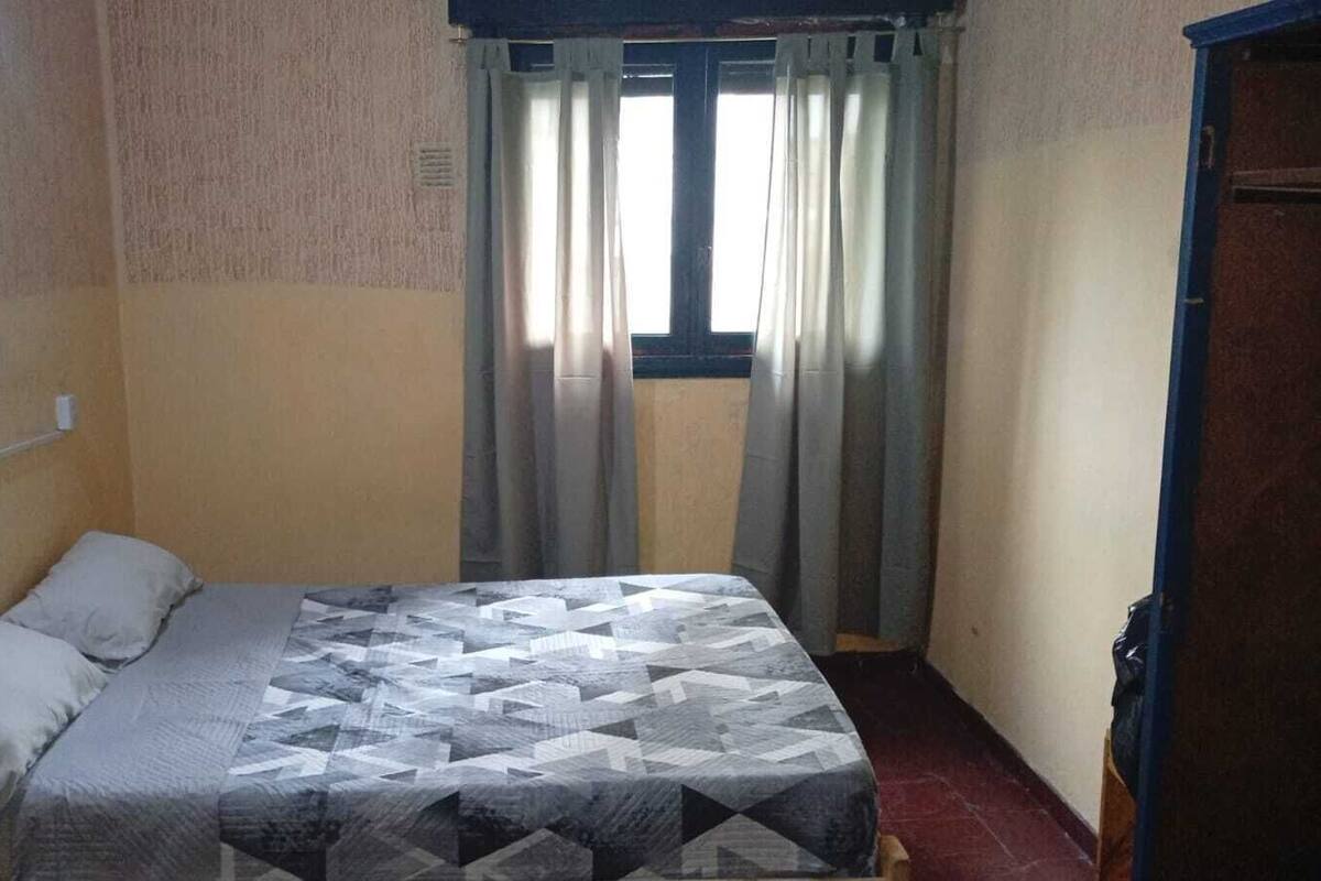 Quarto pequeno com uma cama de solteiro, colcha estampada, janela com cortinas e uma porta à direita.