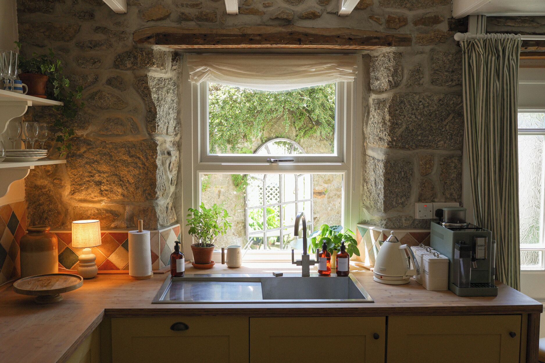 Uma cozinha com parede de pedra, janela com vista para o verde, pia e diversos utensílios de cozinha.