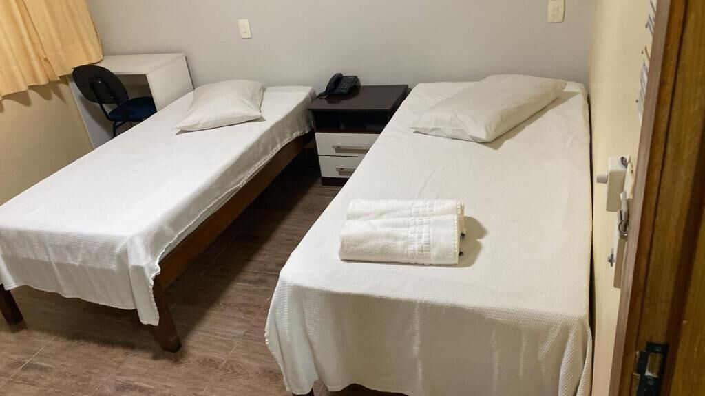 Duas camas de solteiro com lençóis brancos, uma escrivaninha com telefone e uma cadeira.