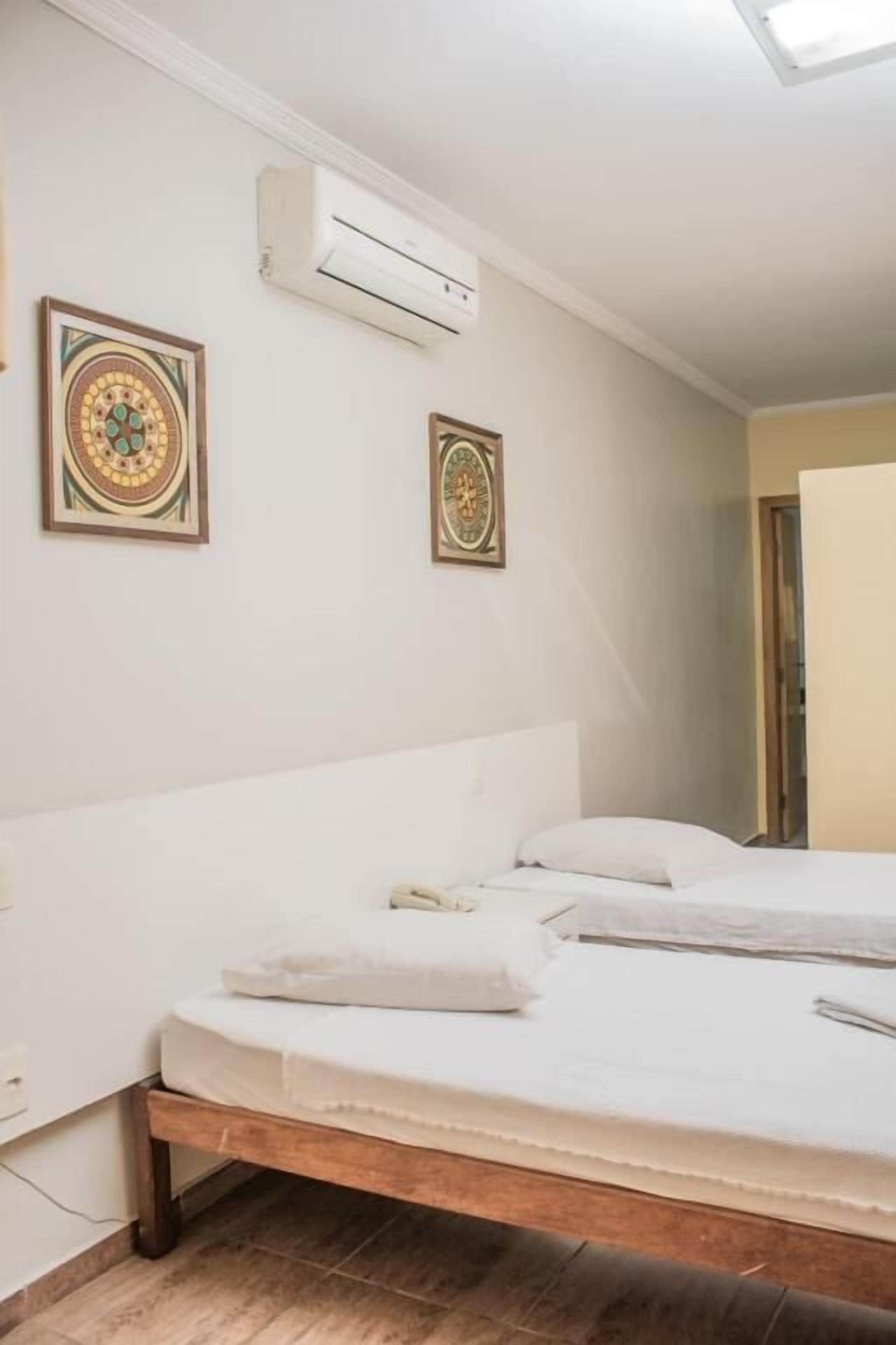 Quarto com cama de madeira, roupa de cama branca, duas unidades de ar condicionado fixadas na parede e dois objetos decorativos emoldurados.