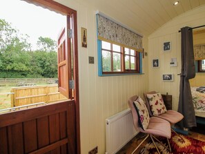 Quarto com porta de madeira que dá para área externa, janela com moldura azul, radiador e cadeira com almofadas florais.