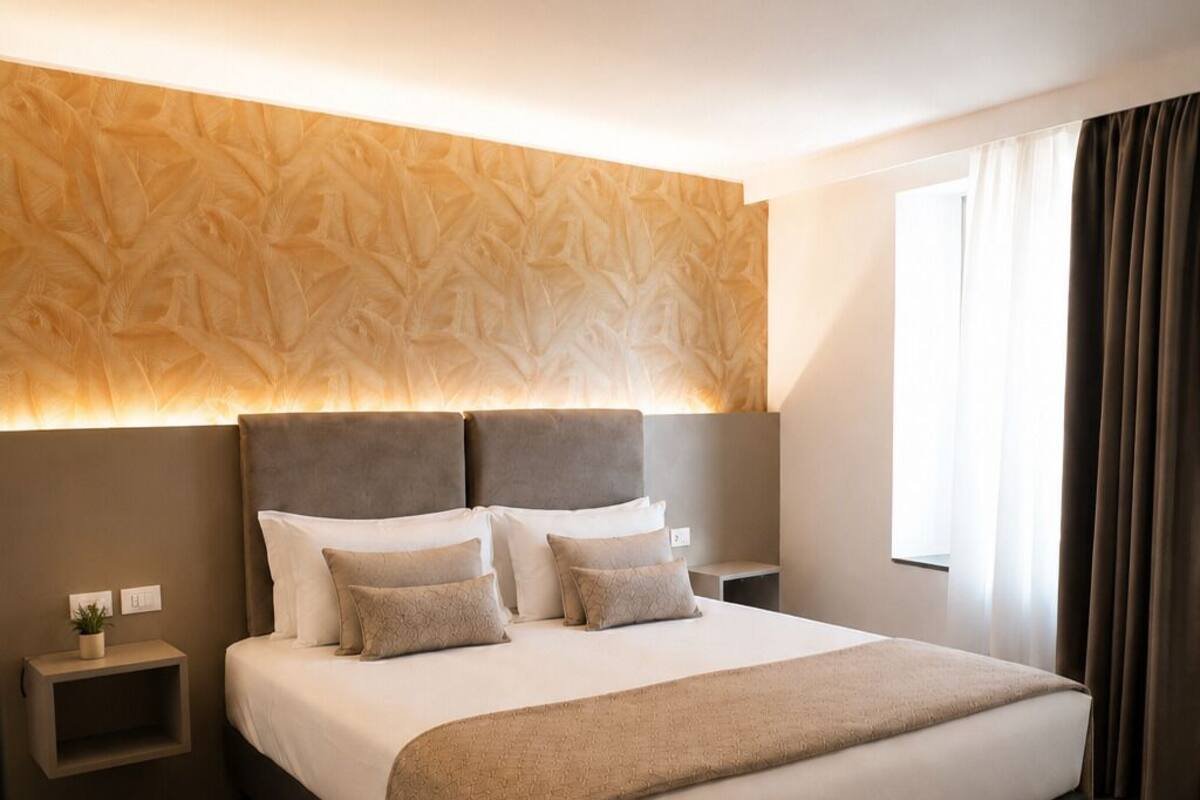 Quarto de hotel moderno com uma cama grande, mesa de cabeceira, luminária na parede e janela com cortinas.