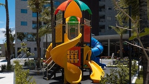 Um parquinho com escorrega e estruturas de escalada, cercado por palmeiras e um edifício moderno.