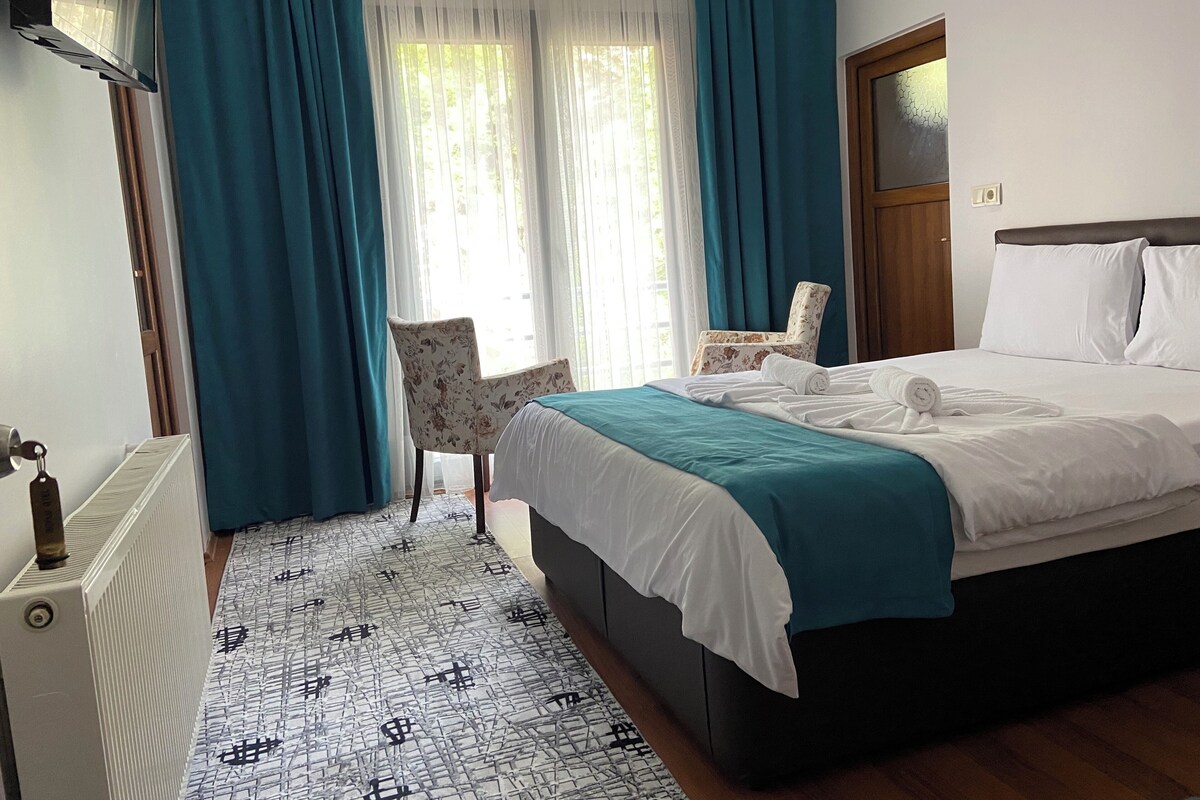 Quarto de hotel com uma cama, duas cadeiras, um radiador e uma janela com cortinas.