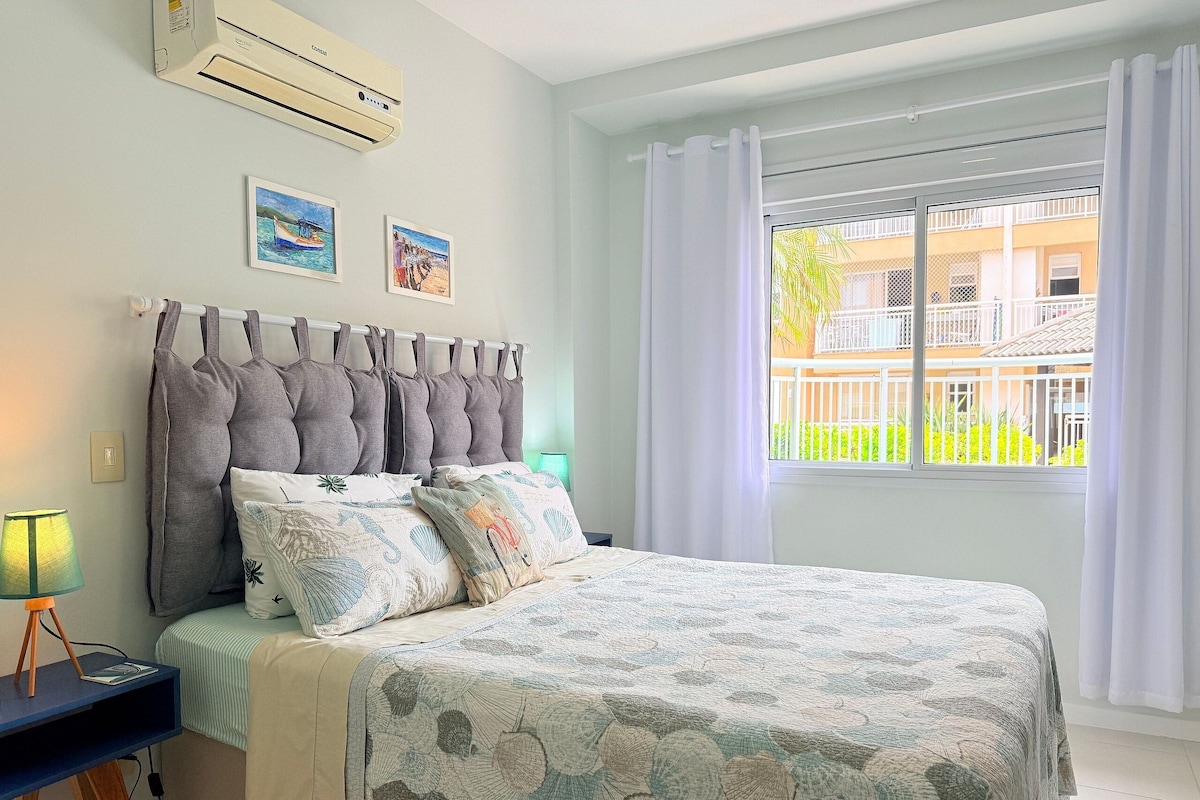 Quarto com cama, ar condicionado, duas pinturas, janela com cortinas e uma luminária.