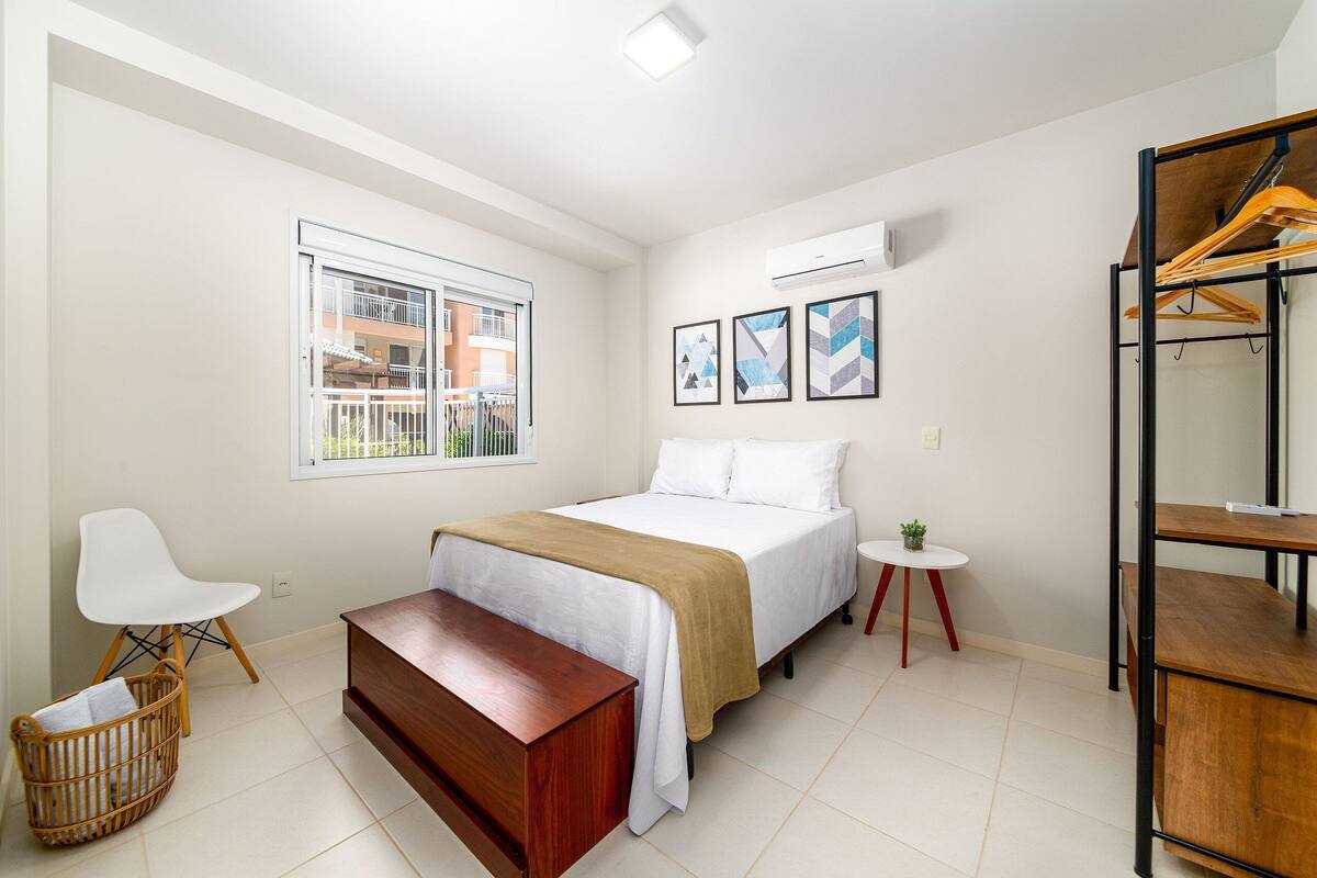 Quarto com cama, um banco, uma cadeira, uma mesinha e um cabideiro.