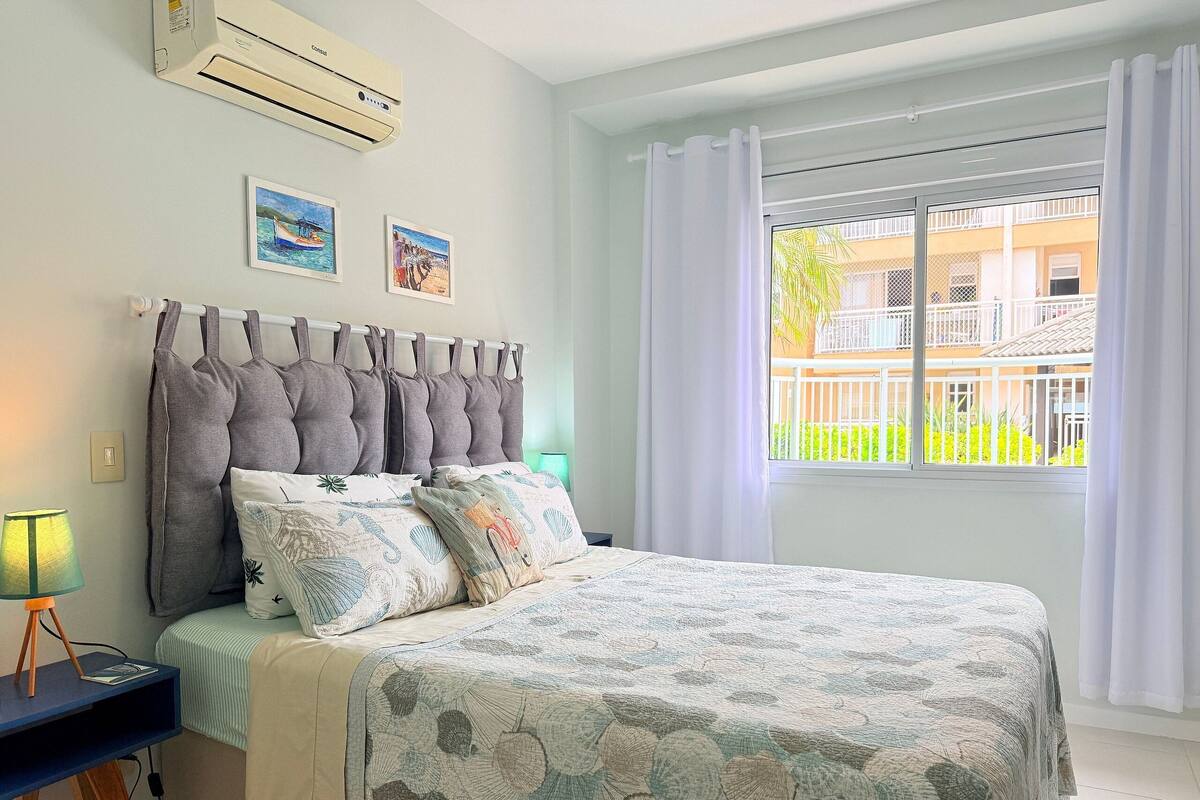 Quarto com cama, ar condicionado, duas pinturas, janela com cortinas e uma luminária.