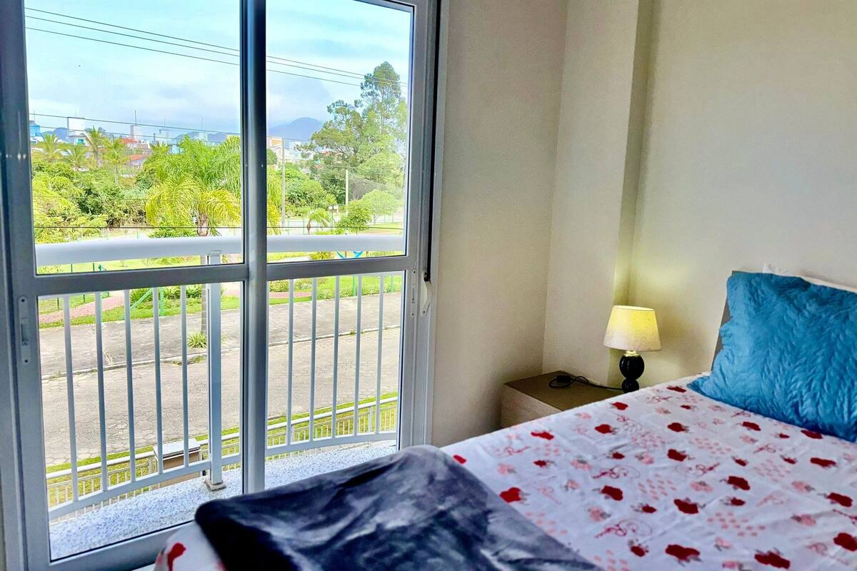Quarto de hotel com uma cama, uma mesa de cabeceira com abajur, janela com vista para a rua e vegetação, e varanda com guarda-corpo.