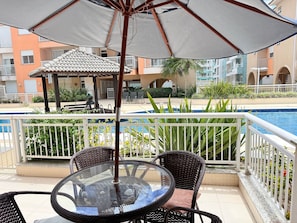 Mesa de vidro com cadeiras de vime, um guarda-sol e vista para a piscina de um edifício com varandas.