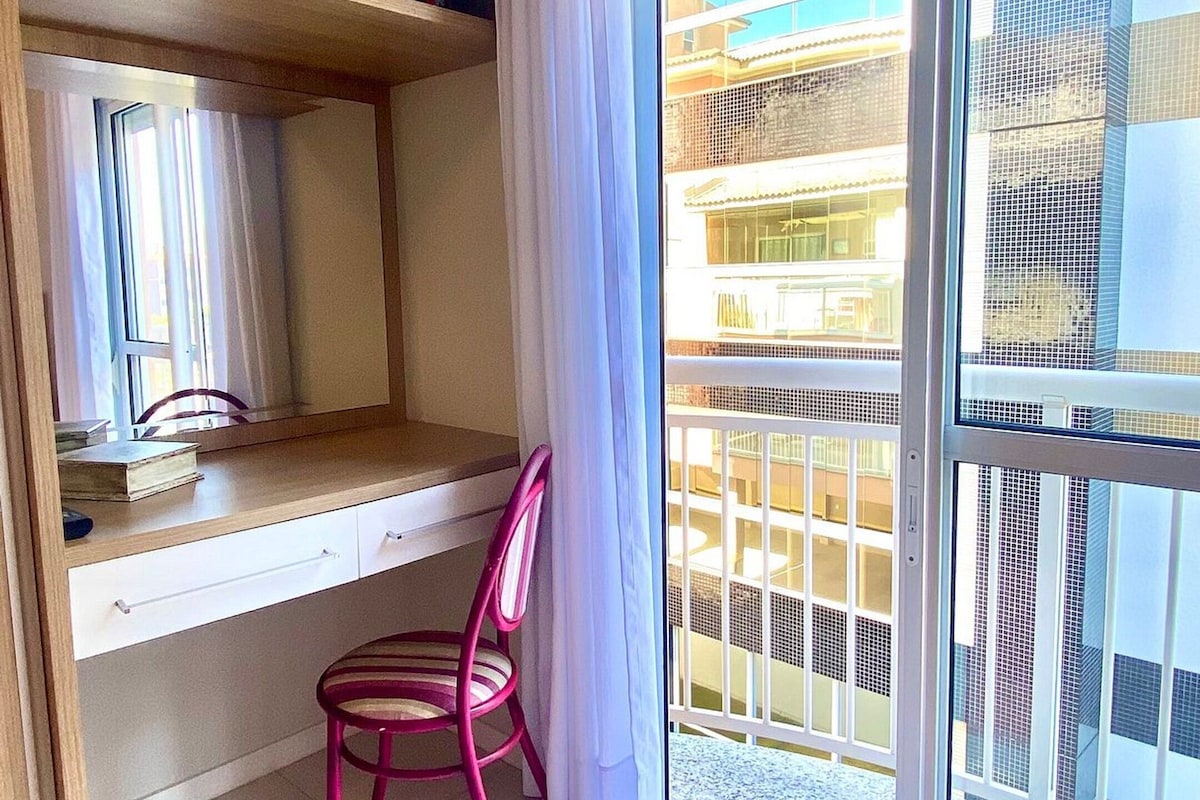 Quarto com cama, televisão em uma estante, uma cadeira e varanda com vista para prédios.