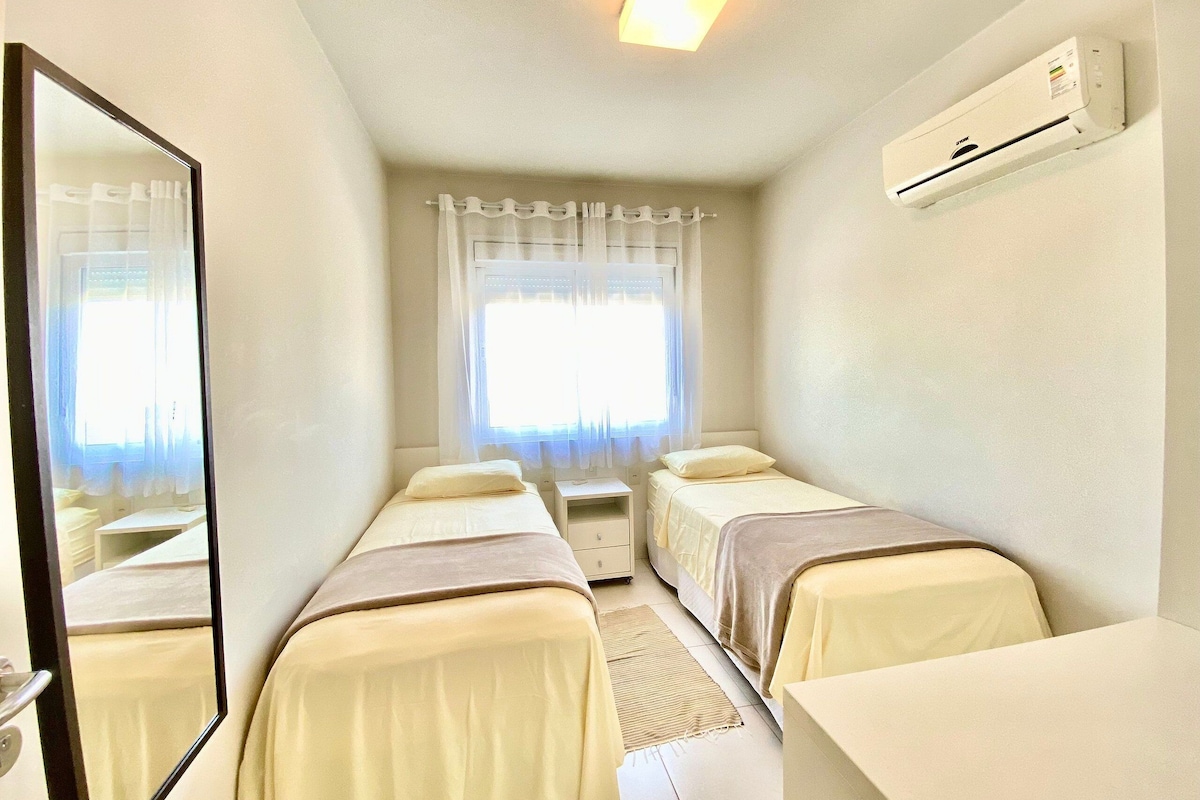 Quarto de hotel com duas camas de solteiro, uma escrivaninha, um espelho, uma janela com cortinas finas e um ar condicionado na parede.