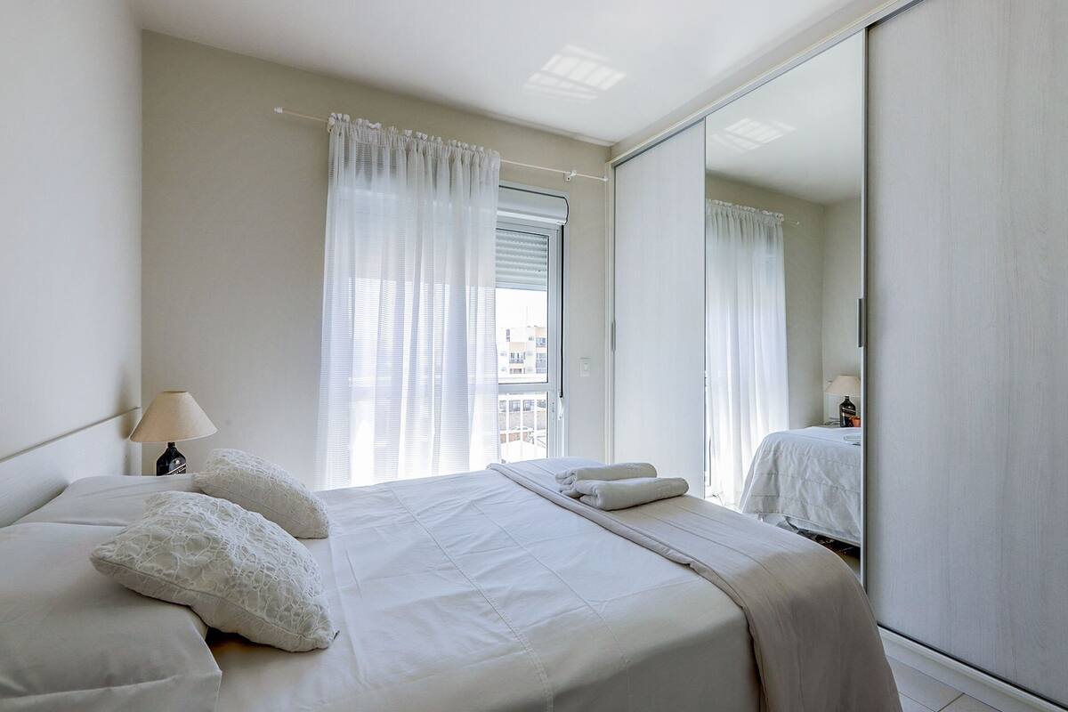 Quarto de hotel com cama, abajur, janela com cortinas e espelho.