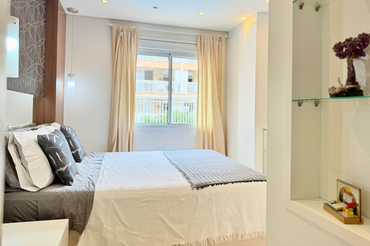 Um quarto com cama, janela com cortinas, mesa de cabeceira com abajur e prateleira de vidro com objetos decorativos.