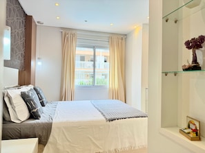 Um quarto com cama, janela com cortinas, mesa de cabeceira com abajur e prateleira de vidro com objetos decorativos.