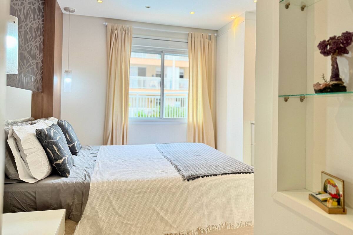 Um quarto com cama, janela com cortinas, mesa de cabeceira com abajur e prateleira de vidro com objetos decorativos.