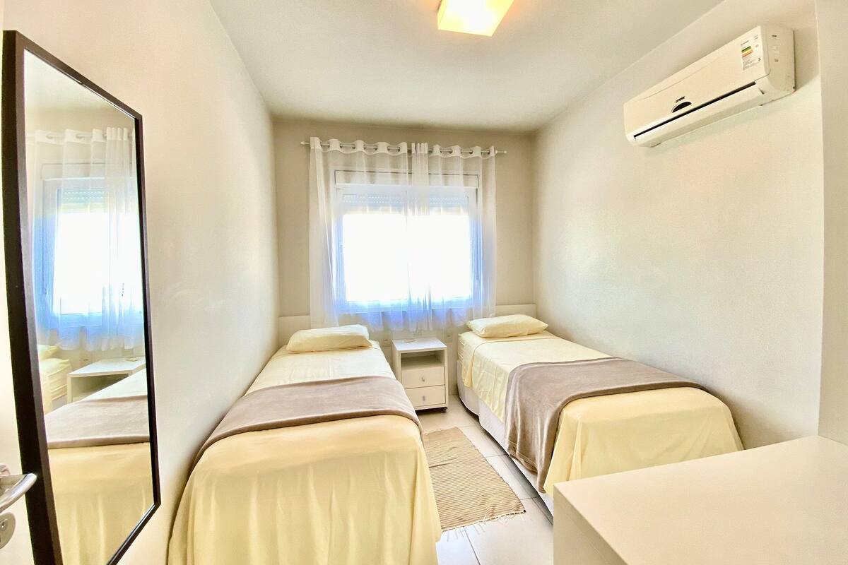Quarto de hotel com duas camas de solteiro, uma escrivaninha, um espelho, uma janela com cortinas finas e um ar condicionado na parede.