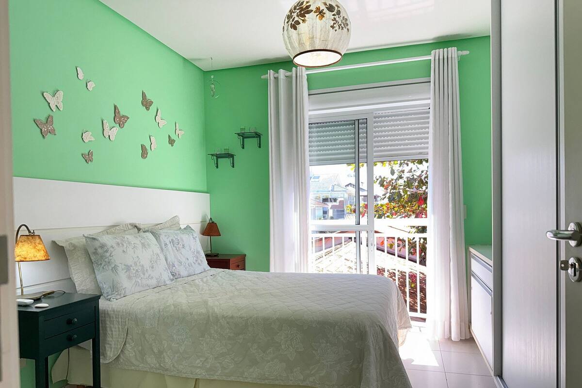 Quarto com cama, mesas de cabeceira, janela com cortinas e vista para o exterior.
