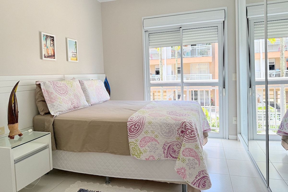 Quarto com cama, mesas de cabeceira, janela com vista para varandas e uma porta.