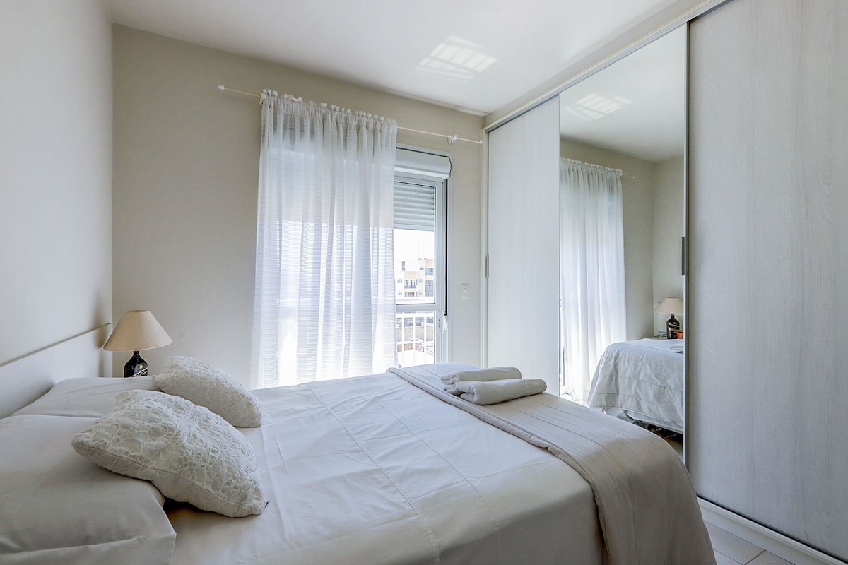 Quarto de hotel com cama, abajur, janela com cortinas e espelho.