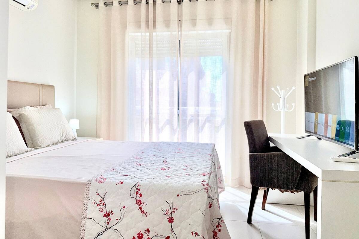 Quarto de hotel com uma cama, uma secretária com televisão e uma janela com cortinas.