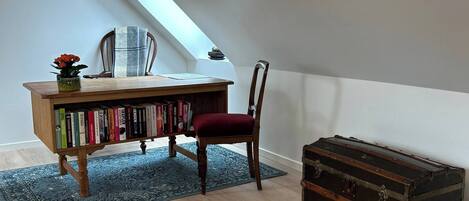 Um quarto com uma estante de madeira, uma cadeira vermelha, uma pequena mesa, um tapete azul e um baú antigo.