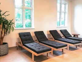 Quarto com quatro espreguiçadeiras de madeira, almofadas pretas, planta em vaso e janelas com vista para o verde.
