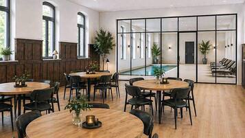 Um hall de hotel com várias mesas redondas, cadeiras, área de piscina e espaço de descanso com espreguiçadeiras.