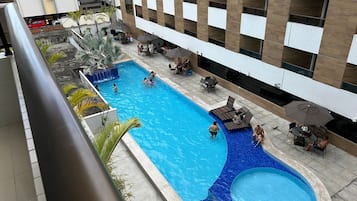 Piscina com várias pistas, rodeada por espreguiçadeiras e guarda-sóis, adjacente a um edifício de design moderno.