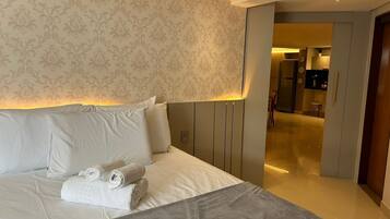 Quarto de hotel com cama, almofadas, cobertor e toalha. Há uma porta que dá para um banheiro e uma área de cozinha.
