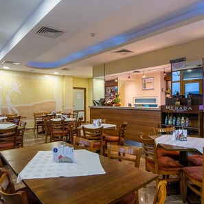 Restaurante com mesas e cadeiras de madeira, área de bar com uma placa indicando 'MENADA W NERY' e um mural na parede.