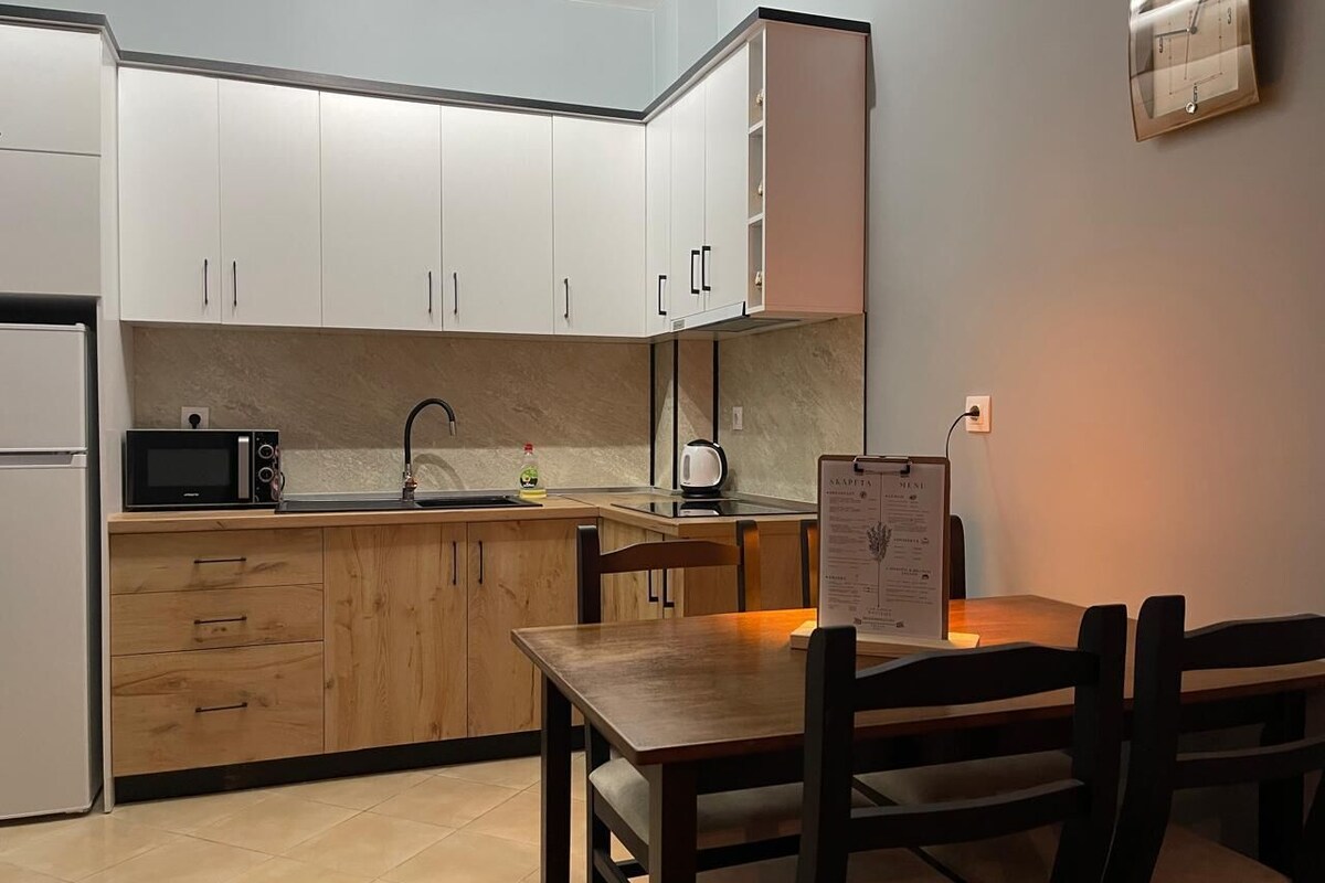 Cozinha compacta com armários brancos, bancada de madeira, micro-ondas, chaleira e mesa de jantar com cadeiras.