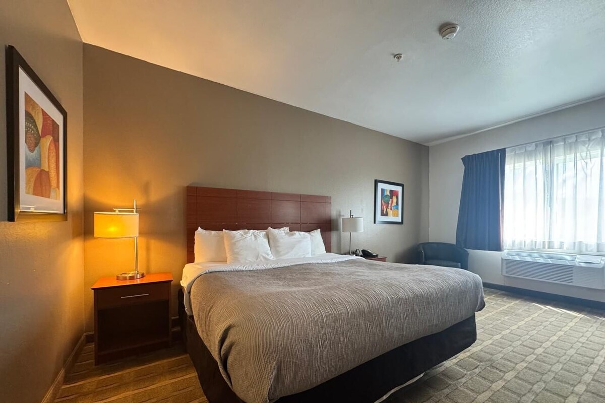 Quarto de hotel com uma cama grande, mesa de cabeceira com abajur, uma cadeira e janela com cortinas.