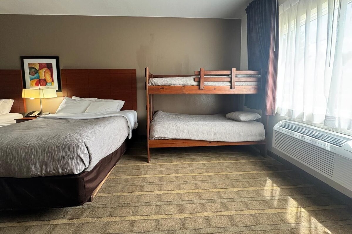Quarto de hotel com duas camas, uma mesa de cabeceira com abajur, um quadro na parede e uma janela com cortinas.