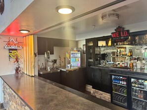 Uma área de bar com um letreiro da Coors Light, um letreiro da Budweiser e uma geladeira cheia de bebidas.