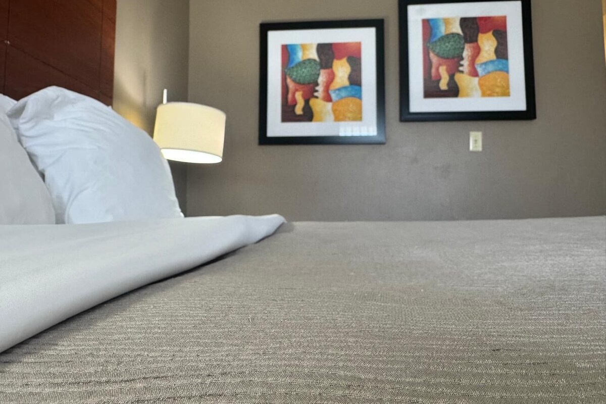 Quarto de hotel com uma cama, uma mesinha de cabeceira com abajur e duas obras de arte emolduradas na parede.
