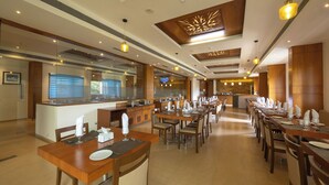 Ampla sala de jantar de hotel, com várias mesas e cadeiras, um buffet central e uma tela de televisão.