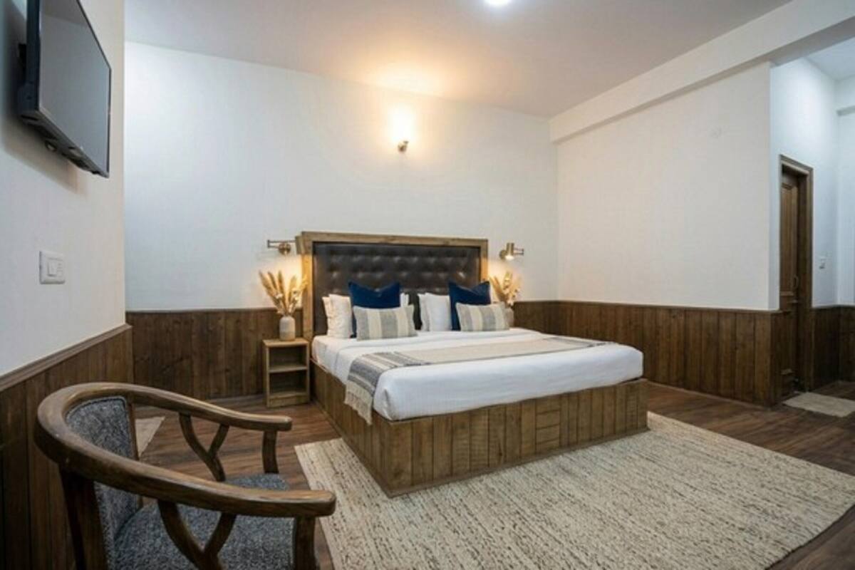 Quarto com cabeceira de madeira, cama com roupa de cama branca, mesa de cabeceira pequena, uma cadeira e uma TV de tela plana na parede.